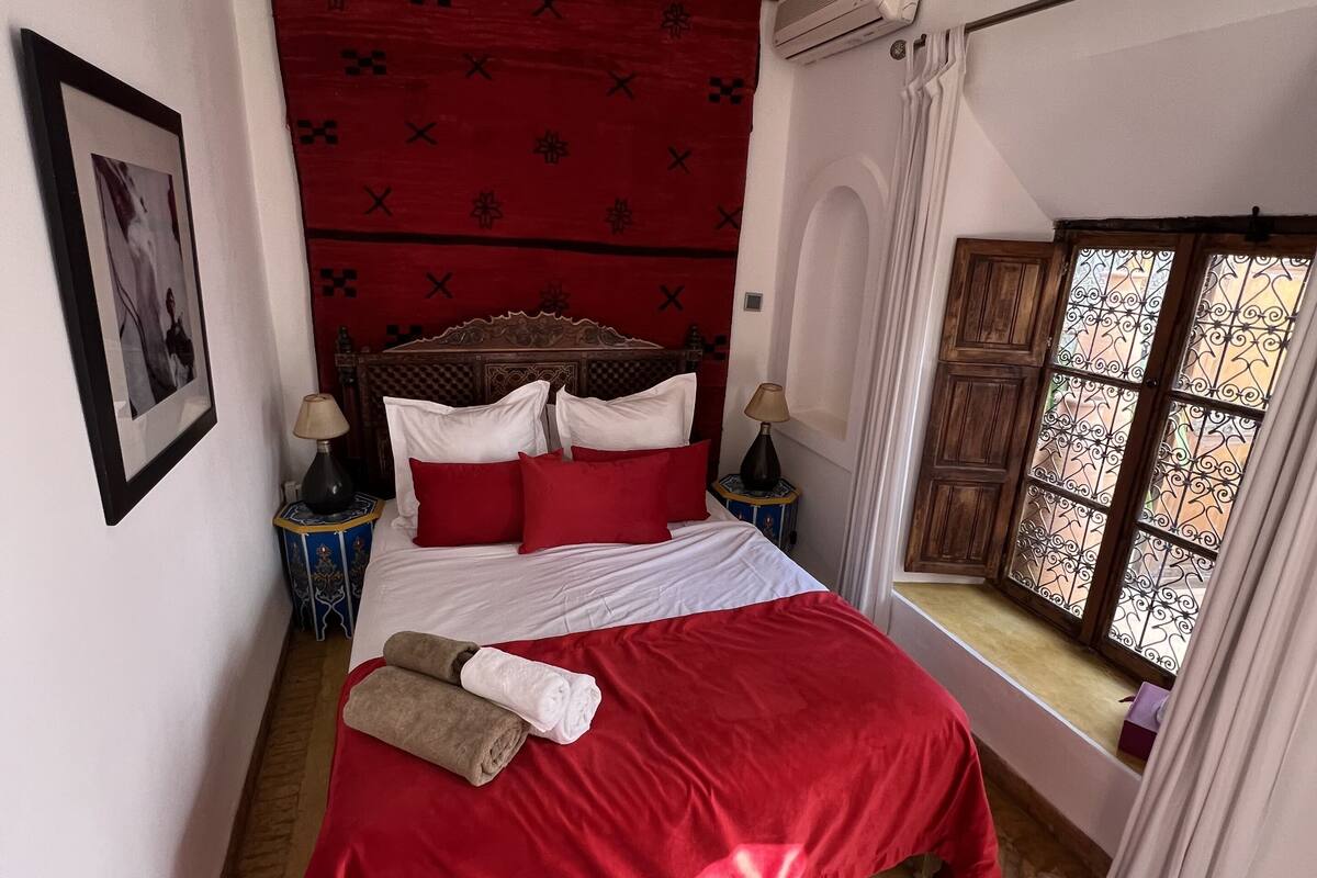 Quarto com cama, cabeceira vermelha, janela com grades de ferro decorativas, tapete estampado e quadro na parede.