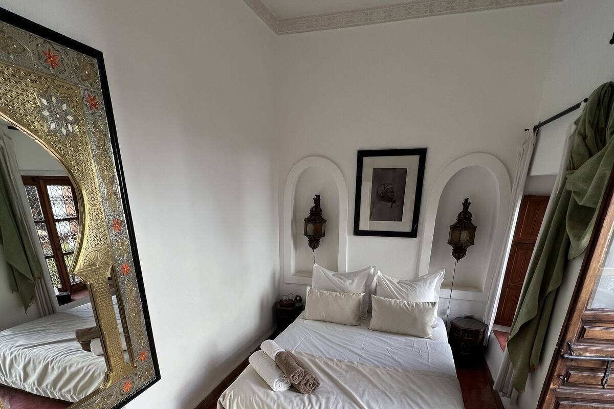 Quarto com uma cama, um espelho grande e ornamentado, duas luminárias fixadas na parede e um quadro emoldurado.