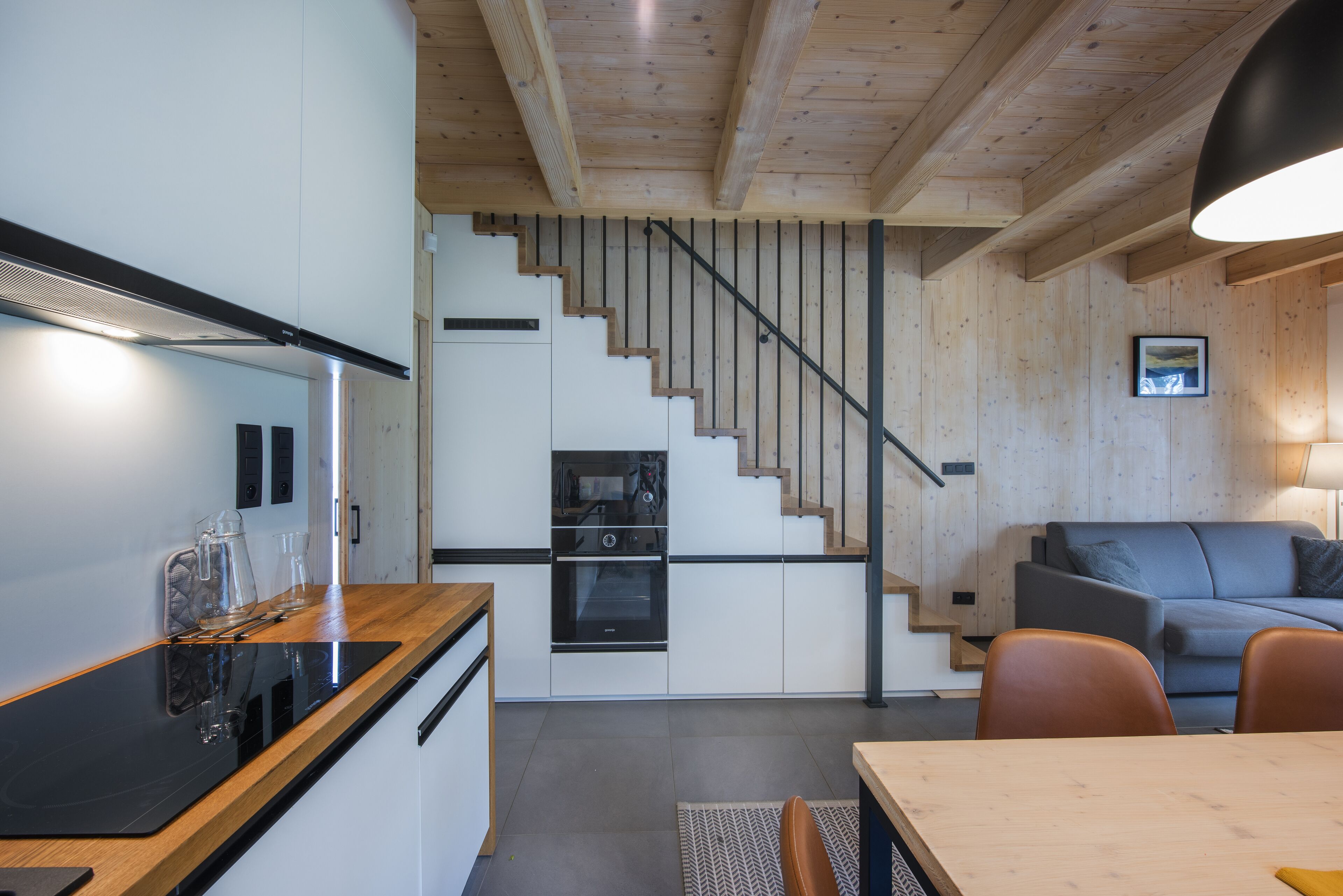 Cozinha moderna com bancada preta, armários de madeira e forno embutido. Escada leva a uma sala de estar com sofá cinza e mesa de centro.