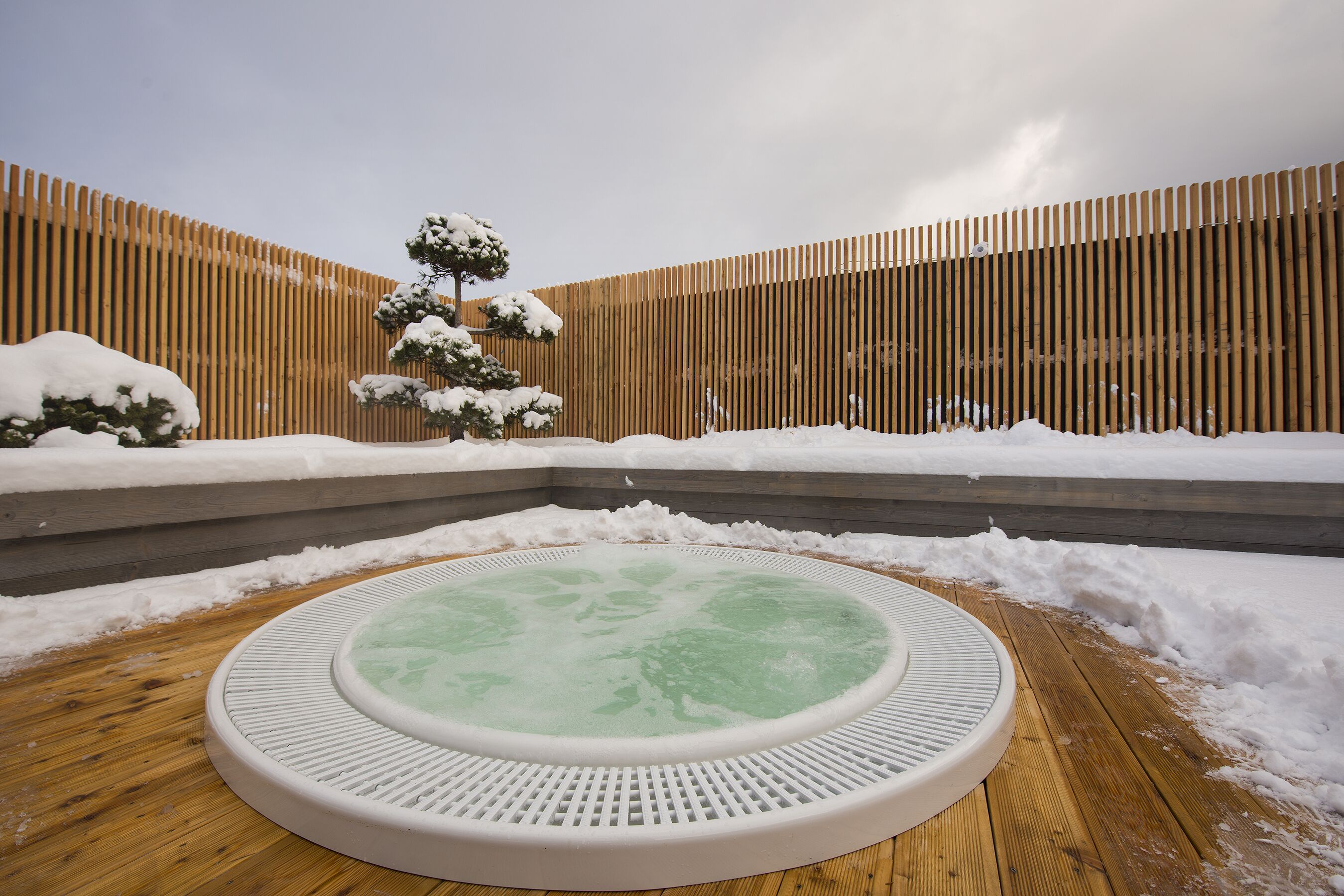 Uma banheira de hidromassagem cercada por neve, com uma cerca de madeira e uma árvore ao fundo.