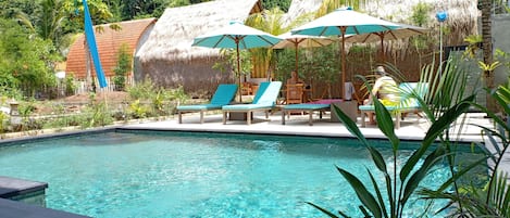 Uma área de piscina com espreguiçadeiras e guarda-sóis, cercada por edifícios com telhado de palha e vegetação exuberante.