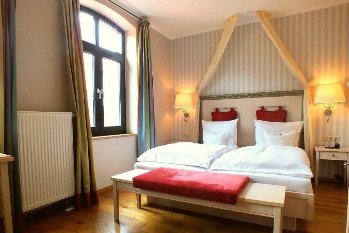 Quarto com cama, mesas de cabeceira, janela com cortinas e radiador.