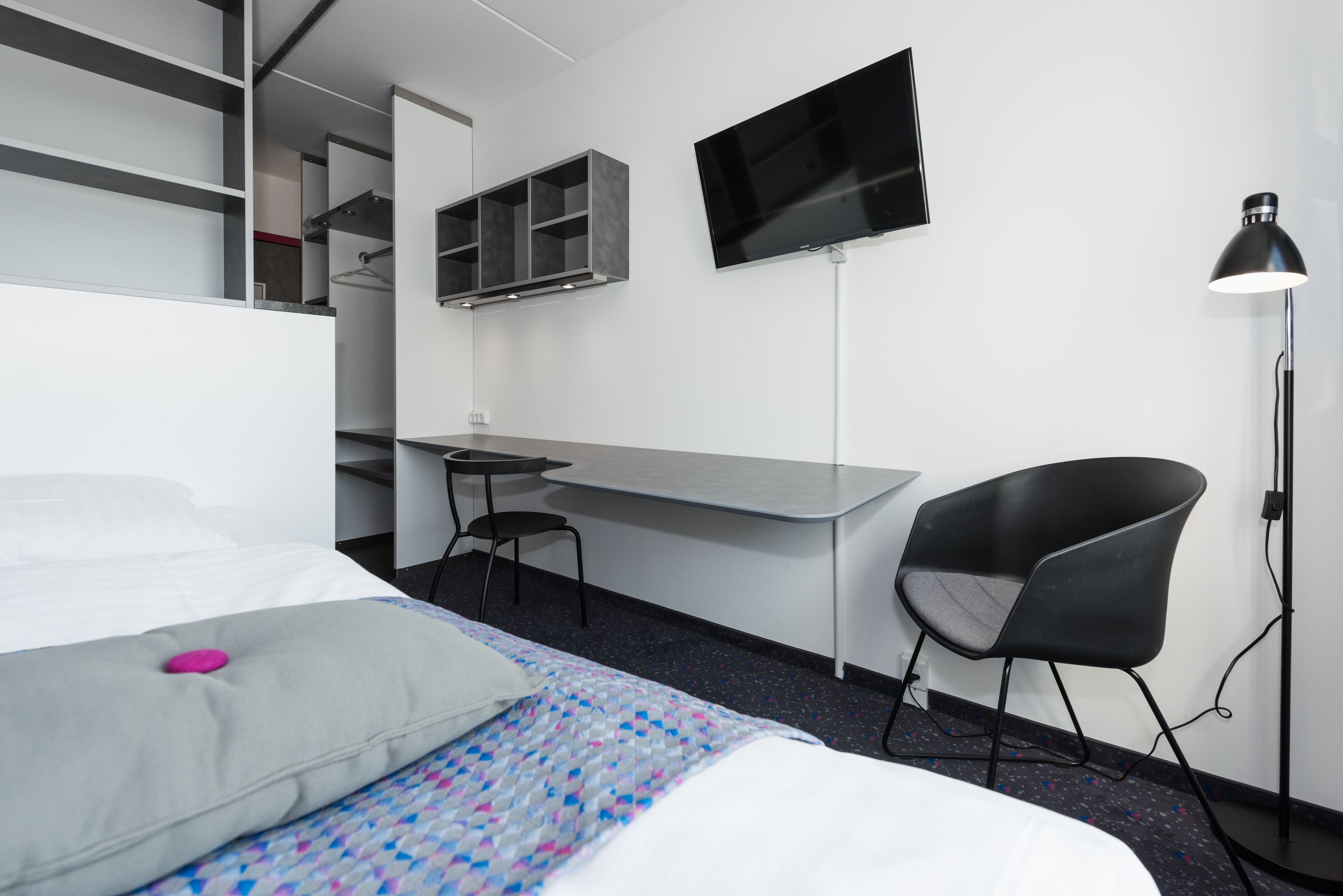 Quarto de hotel com cama, escrivaninha, cadeira, televisão e abajur.