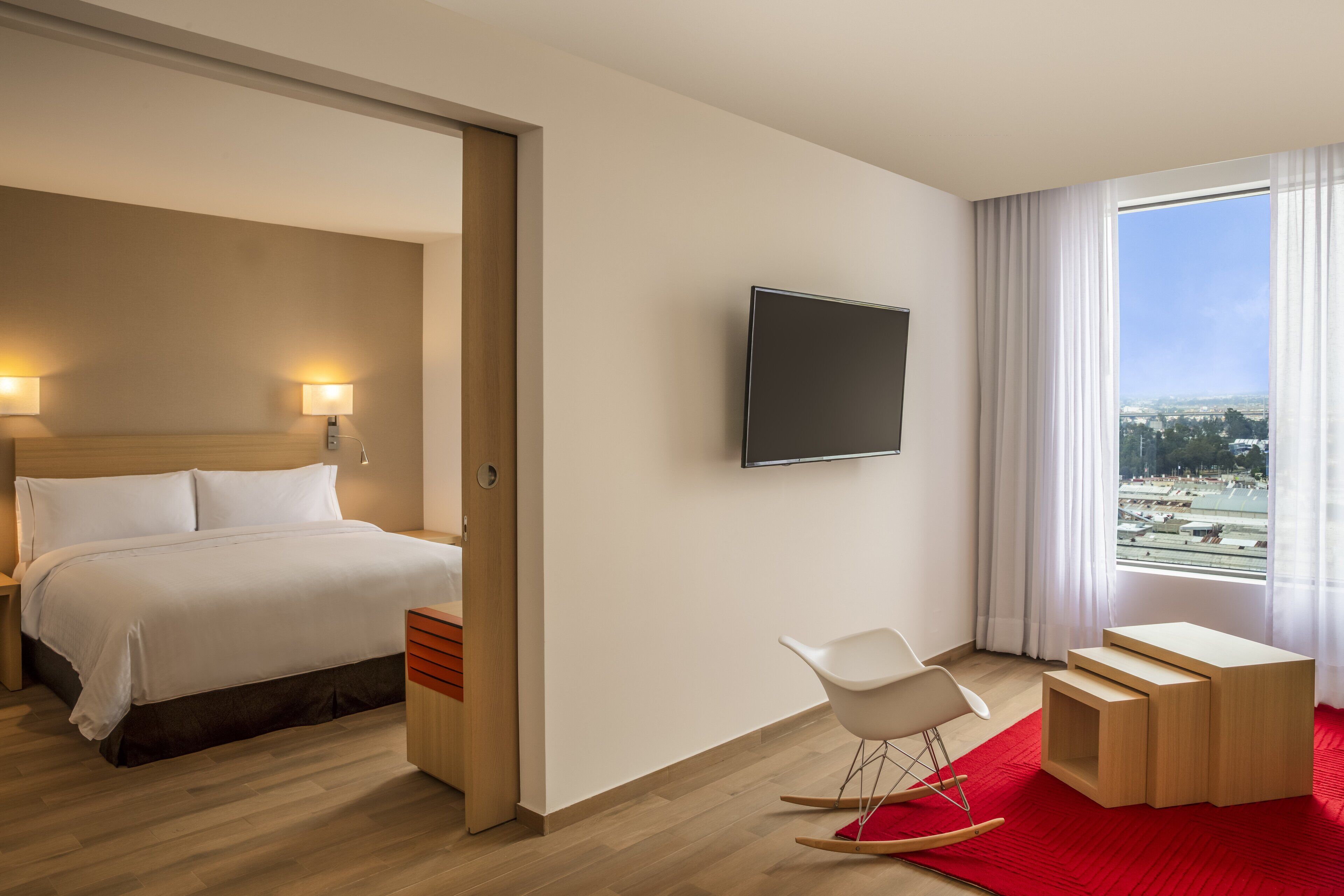 Quarto de hotel moderno com cama, televisão, cadeira de balanço e vista da cidade.