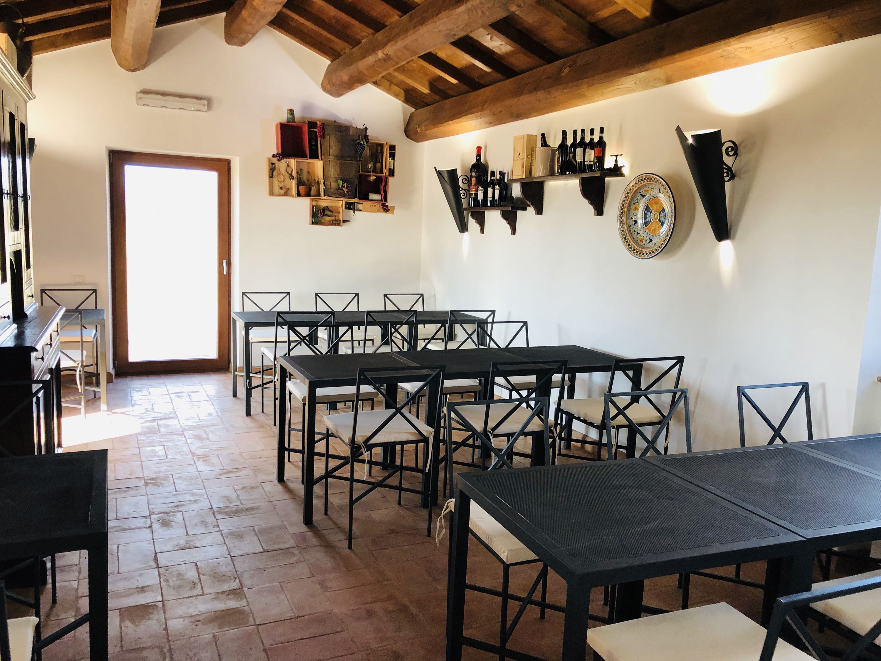 Uma área de refeições com várias mesas e cadeiras, vigas de madeira no teto e garrafas de vinho expostas em prateleiras.
