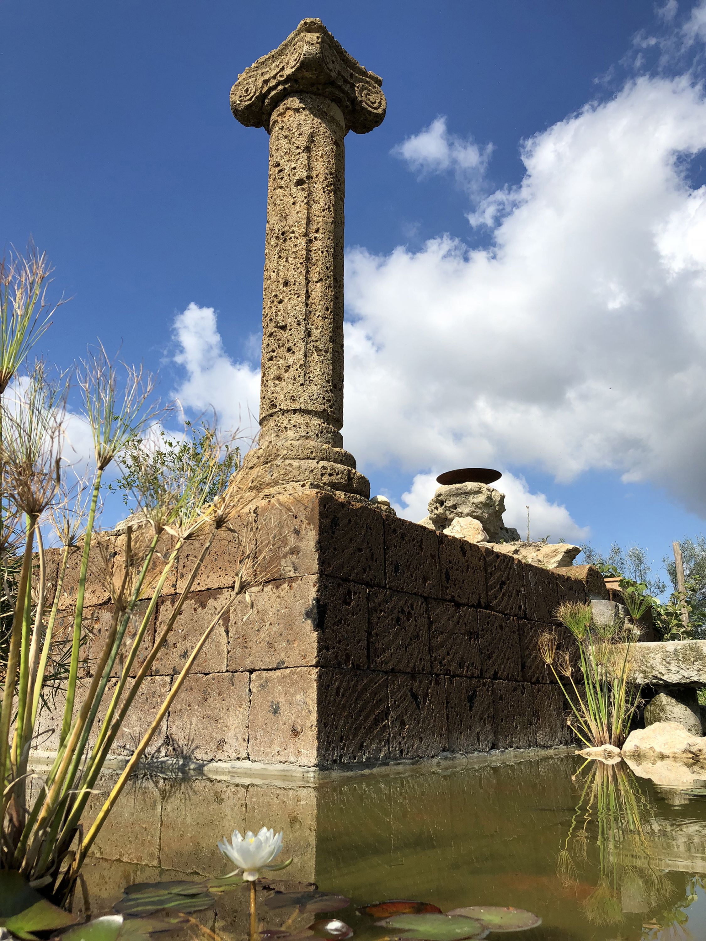 Um pilar de pedra com capitel no topo, rodeado por um lago com folhas de nenúfar e vitórias-régias.