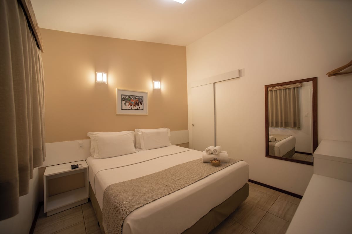 Quarto de hotel com cama, mesa de cabeceira, luminárias na parede, espelho e um quadro.