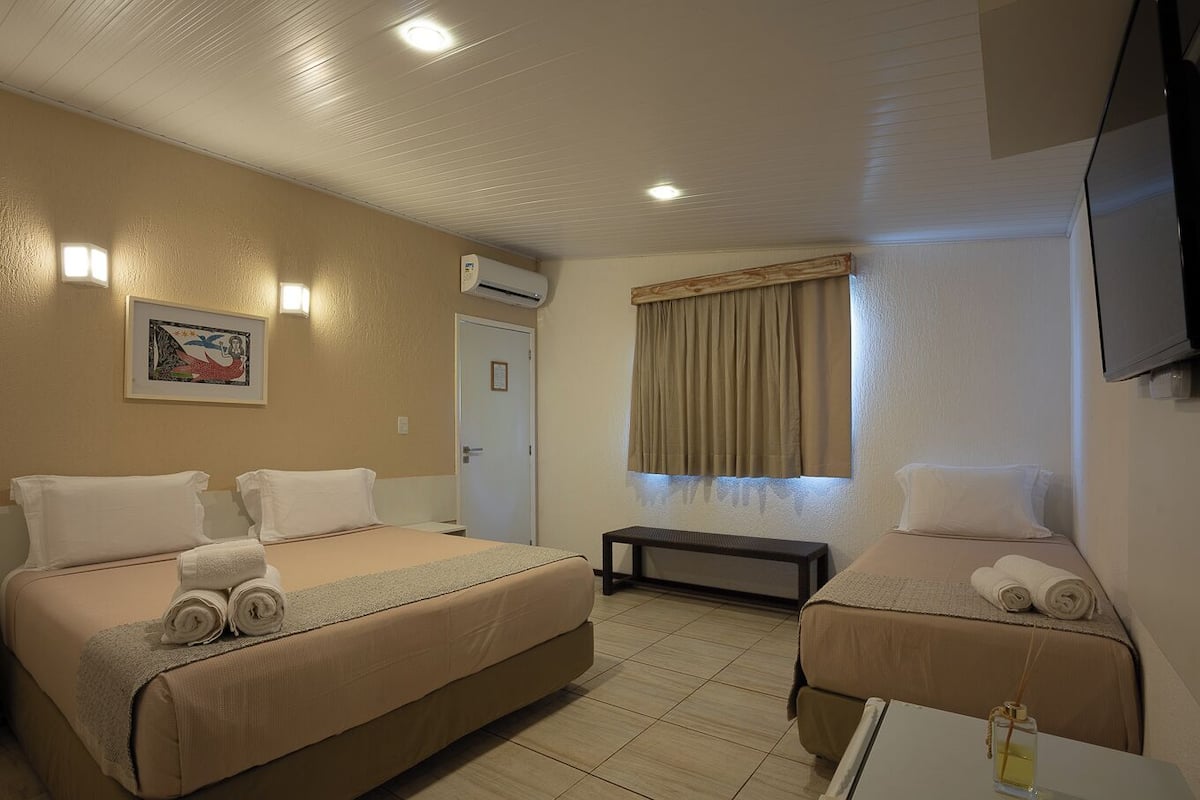 Quarto de hotel com duas camas, uma televisão, aparelho de ar condicionado, um quadro, uma janela com cortinas e uma pequena mesa com uma garrafa.