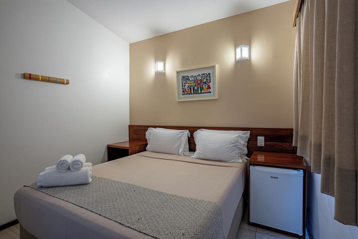 Quarto de hotel com cama, mesinha de cabeceira, luminária na parede, quadro e cortina.