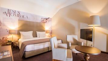 Quarto de hotel com cama, duas mesas de cabeceira, televisão, cadeira e luminária.