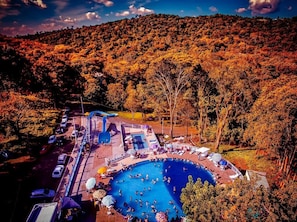 Piscina cercada por cadeiras de praia e guarda-sóis, com uma encosta arborizada ao fundo.