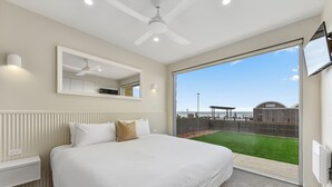 Quarto com cama grande, ventilador de teto, TV na parede e porta de vidro deslizante para uma varanda com vista para a praia.