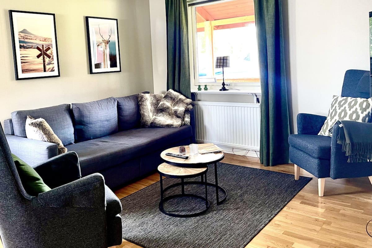 Uma sala de estar com um sofá azul escuro, uma mesa de centro redonda e uma poltrona azul. Há duas imagens emolduradas na parede, uma janela com cortinas e um radiador sob a janela.