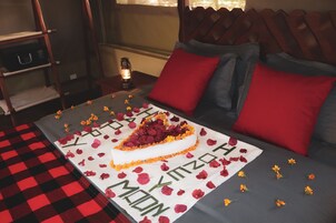 Cama com um bolo em formato de coração sobre uma toalha de mesa com a palavra "LOVE", rodeada por pétalas de rosa.