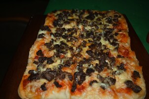 Pizza retangular com massa grossa, coberta com queijo derretido e pedaços de linguiça.