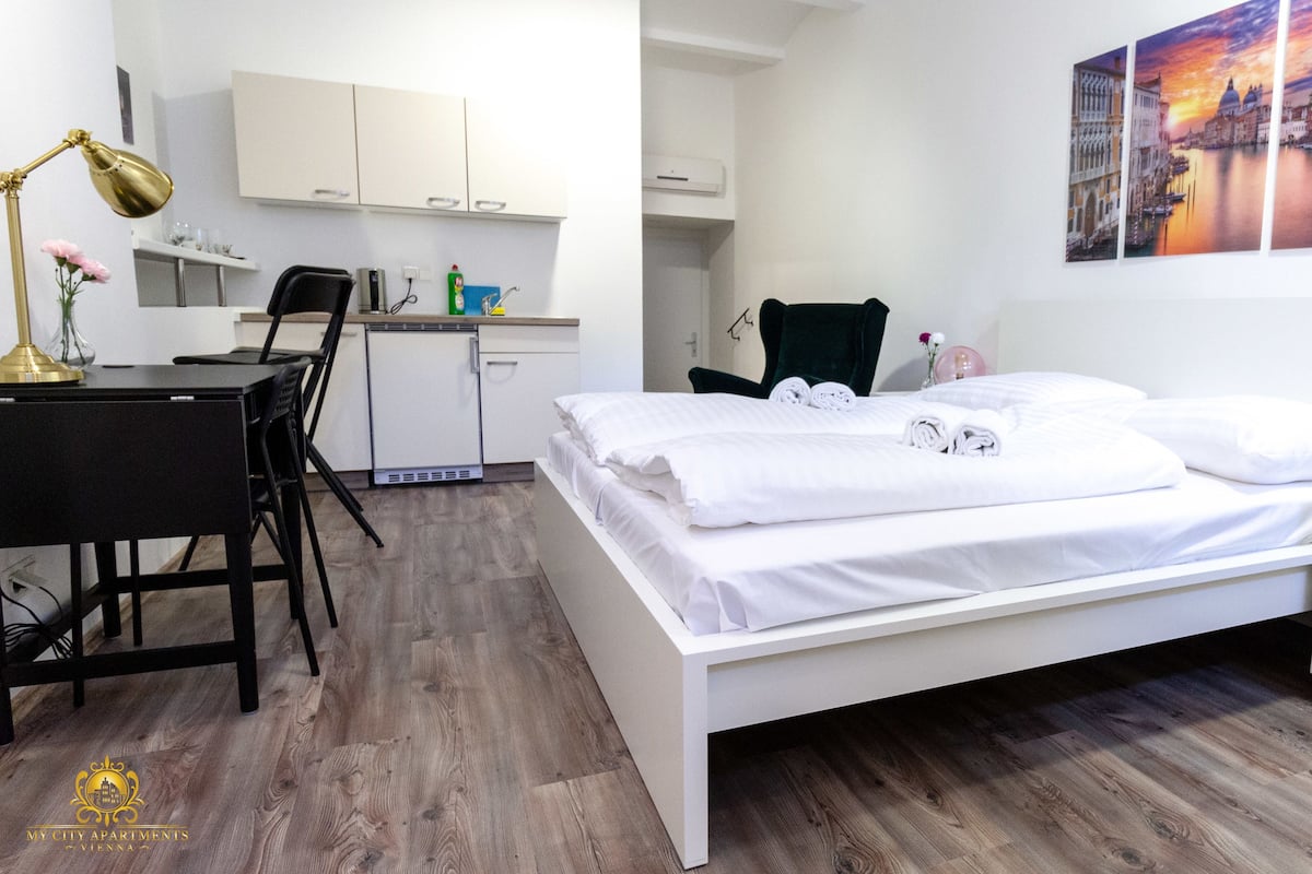 Quarto de hotel com cama, escrivaninha, cadeira, mesa pequena, mesa de cabeceira, luminária e um quadro na parede.