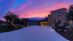 Um edifício moderno com um logotipo 'RO' em destaque, entrada pavimentada e um jardim ajardinado com árvores e arbustos.
