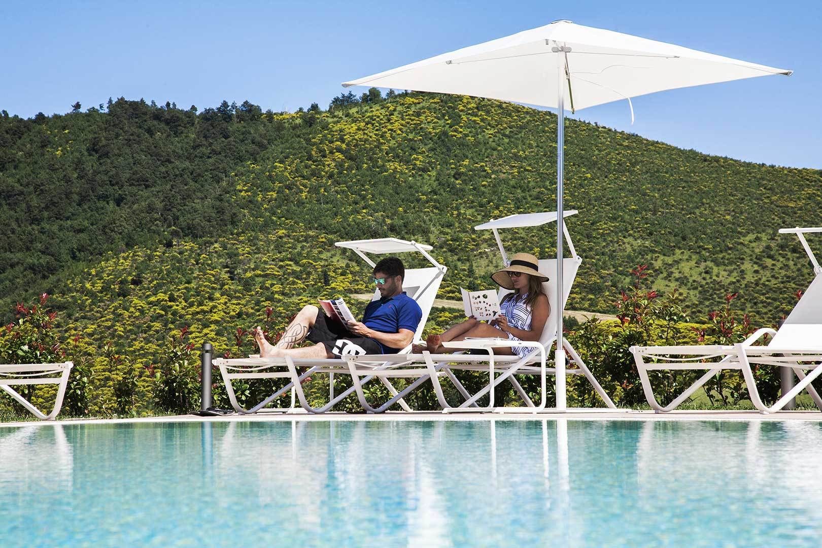 Duas pessoas relaxando perto de uma piscina com espreguiçadeiras brancas e um guarda-sol branco.