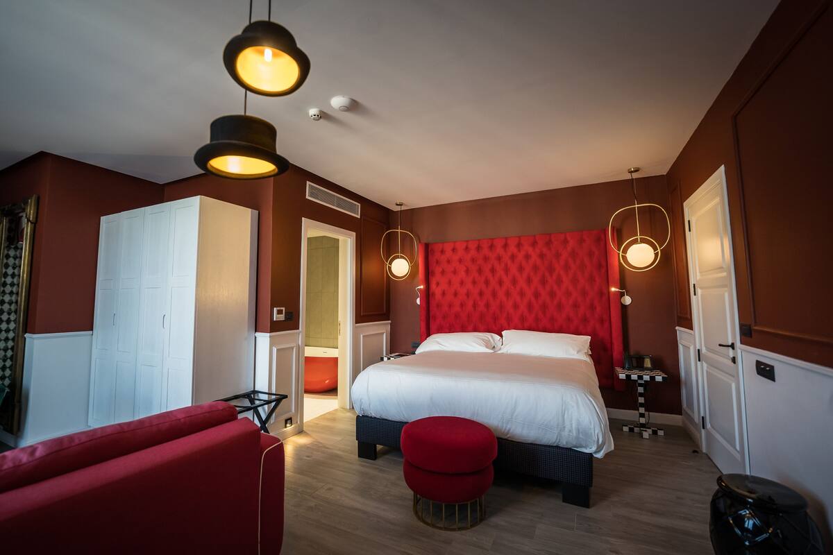 Quarto de hotel moderno com uma cama grande, cabeceira estofada em vermelho e uma poltrona vermelha.