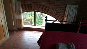 Quarto com colcha vermelha, vista do jardim pela janela e ventilador de teto.