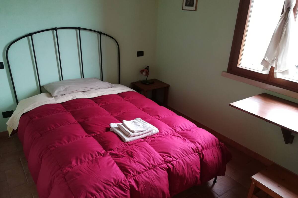 Cama com colcha vermelha, travesseiro branco e toalha dobrada. Pequeno criado-mudo de madeira com abajur ao lado da cama.
