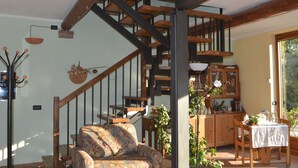 Uma sala de estar ampla com escada de madeira, sofá floral, mesa de jantar com cadeiras e área de cozinha com armários de madeira e plantas em vasos.