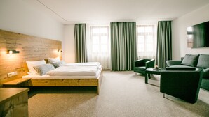 Quarto de hotel moderno com cabeceira de madeira, cama, sofá verde, mesa pequena e televisão montada na parede.