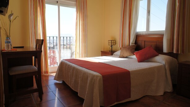 Quarto de hotel com cama, escrivaninha, cadeira, mesa de cabeceira e vista para a cidade pela janela.
