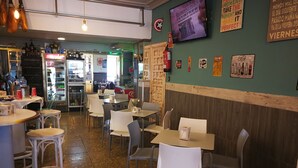 Interior de restaurante com mesas, cadeiras, área de bar e uma televisão exibindo um programa.