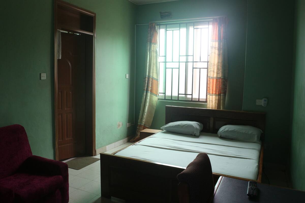 Um quarto pequeno e simples, com uma cama de solteiro, uma cadeira e uma janela com cortinas.