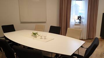 Uma sala de conferências com mesa branca, cadeiras pretas, quadro branco e janela com cortinas.