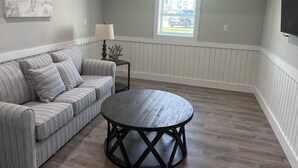 Um quarto com um sofá listrado, uma mesa de madeira redonda, um abajur, uma janela com vista para um carro e um quadro na parede.