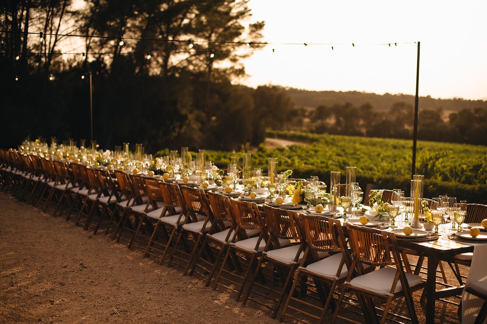 Mesa longa posta ao ar livre com cadeiras, luzes de festa e vegetação ao fundo.