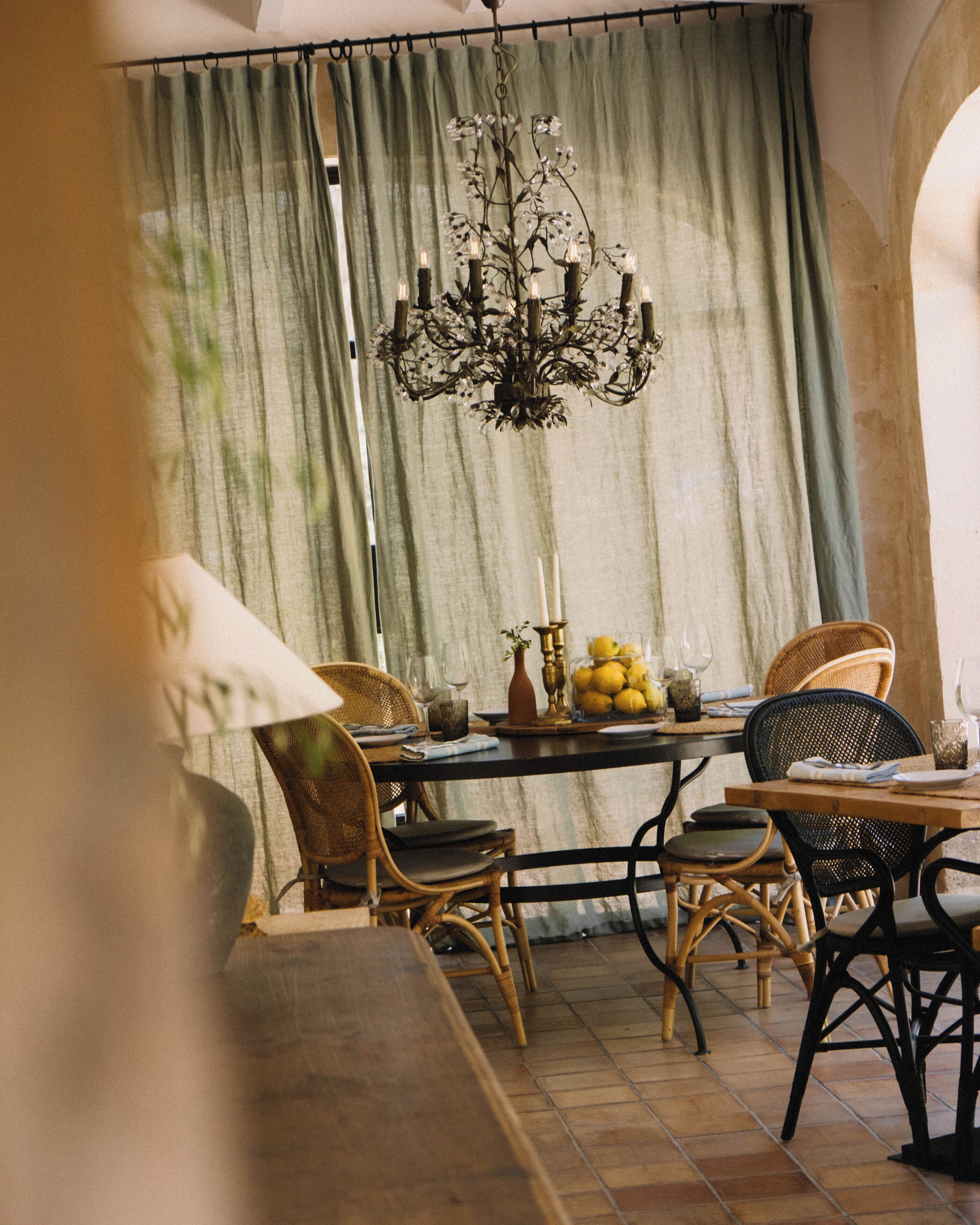 Uma sala de jantar com uma mesa posta para dois, rodeada por cadeiras de vime, um lustre e um painel de cortinas.