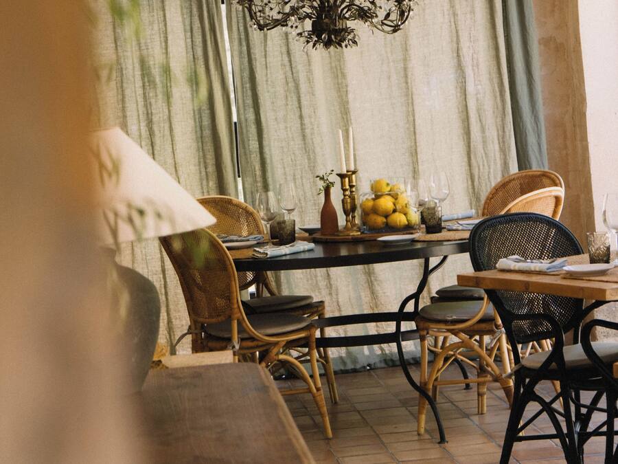 Uma sala de jantar com uma mesa posta para dois, rodeada por cadeiras de vime, um lustre e um painel de cortinas.