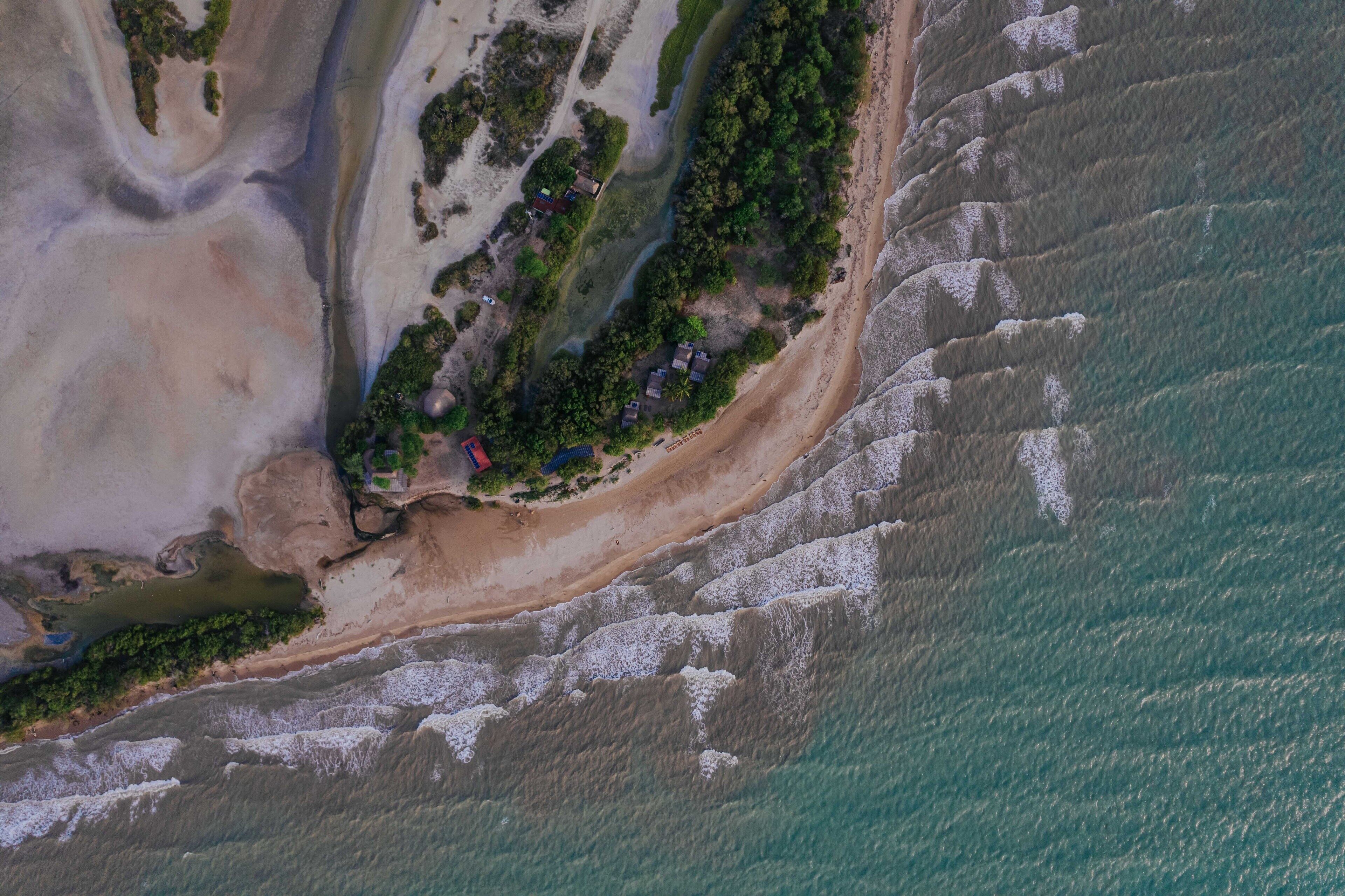 Vista aérea de uma área costeira com praia, rio e um pequeno povoado com edifícios e vegetação.