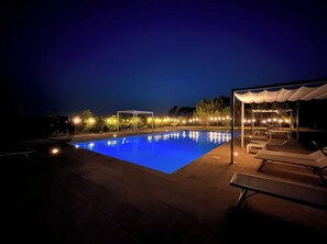 Piscina à noite com superfície azul cristalina, rodeada por espreguiçadeiras e uma área de estar coberta.