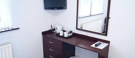 Quarto de hotel com uma escrivaninha de madeira escura, televisão, espelho, radiador, lixeira e um pequeno banquinho.