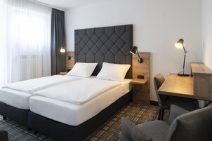 Quarto de hotel com uma cama de casal, duas mesas de cabeceira com abajures, uma escrivaninha de madeira e uma cadeira.