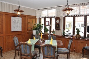 Uma área de refeições com paredes revestidas de madeira, uma janela grande com cortinas de renda, e uma mesa posta para quatro com toalha e guardanapos verdes.