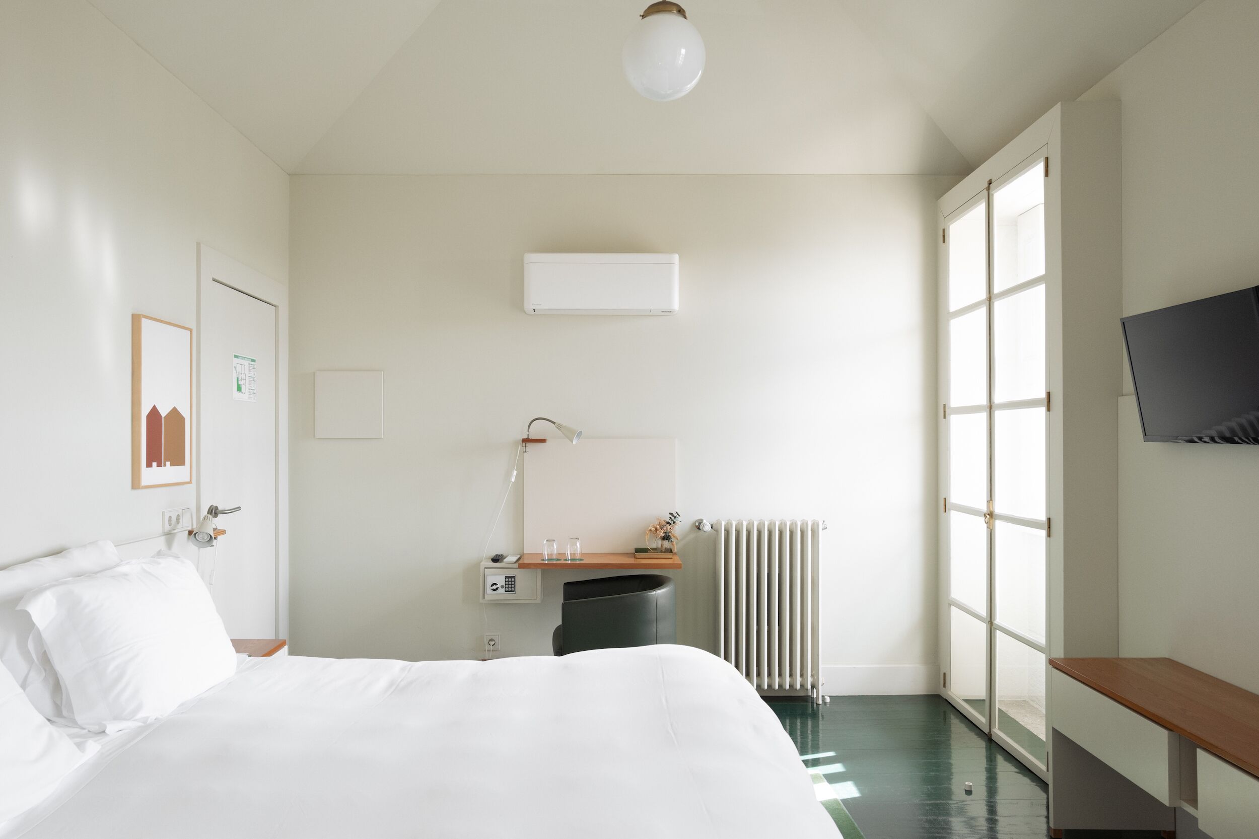 Quarto de hotel com cama, mesa de cabeceira, escrivaninha, cadeira, radiador, televisão e janelas amplas.