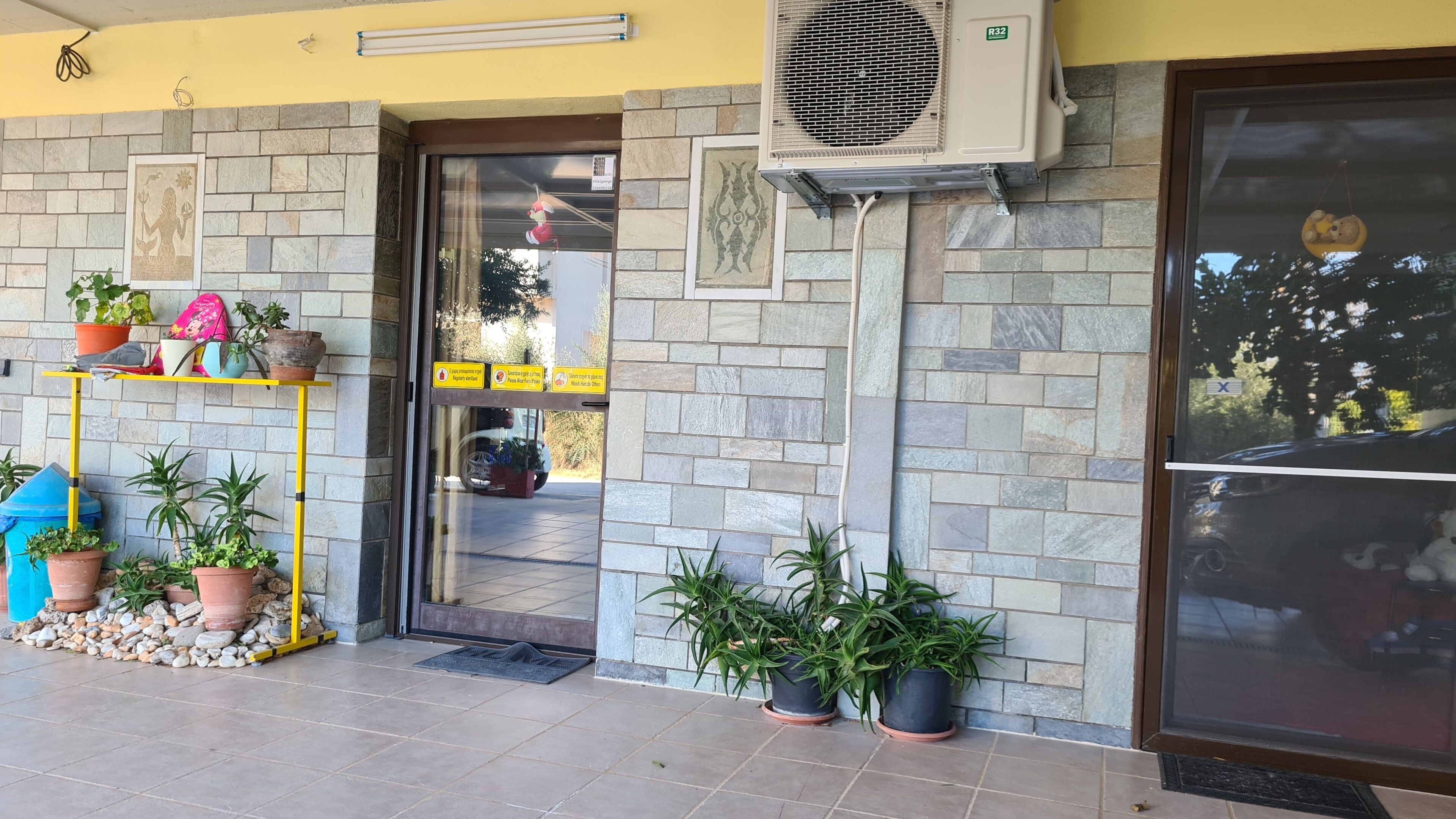 Entrada de um edifício com paredes de pedra, uma porta amarela e plantas em vasos.