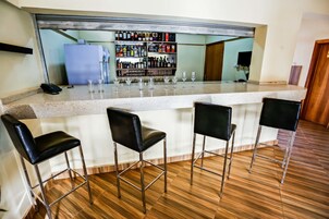 Uma área de bar com balcão, três banquetas pretas e uma estante com diversos copos e garrafas.