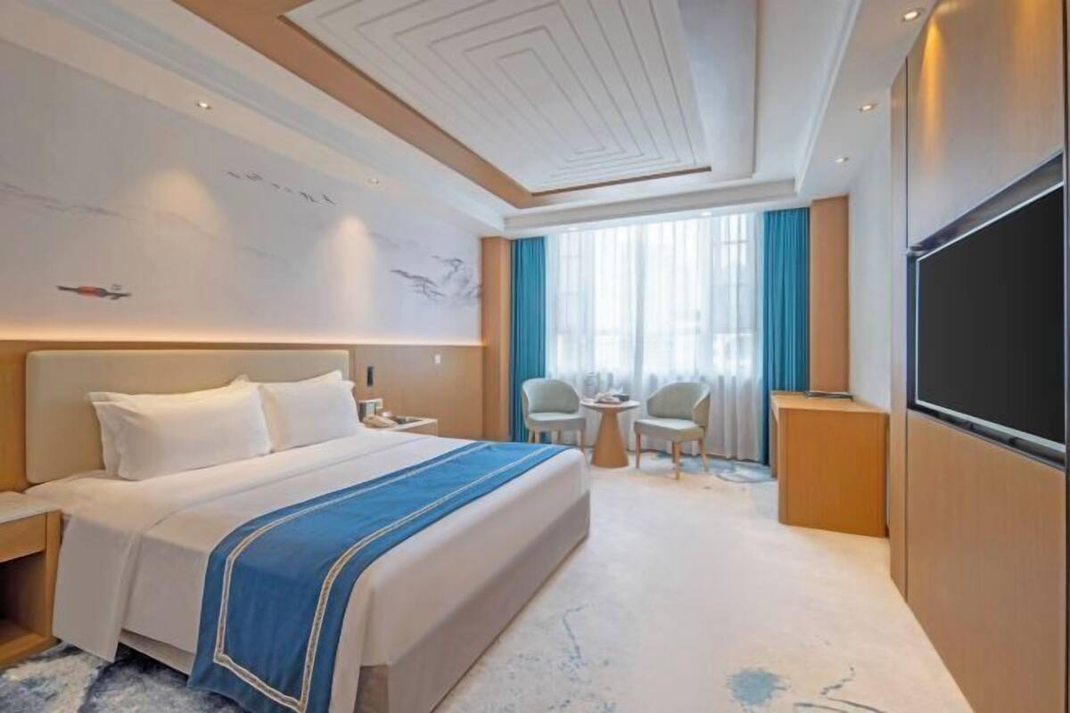 Quarto de hotel com uma cama grande, uma escrivaninha, uma cadeira, uma televisão e uma janela com cortinas.