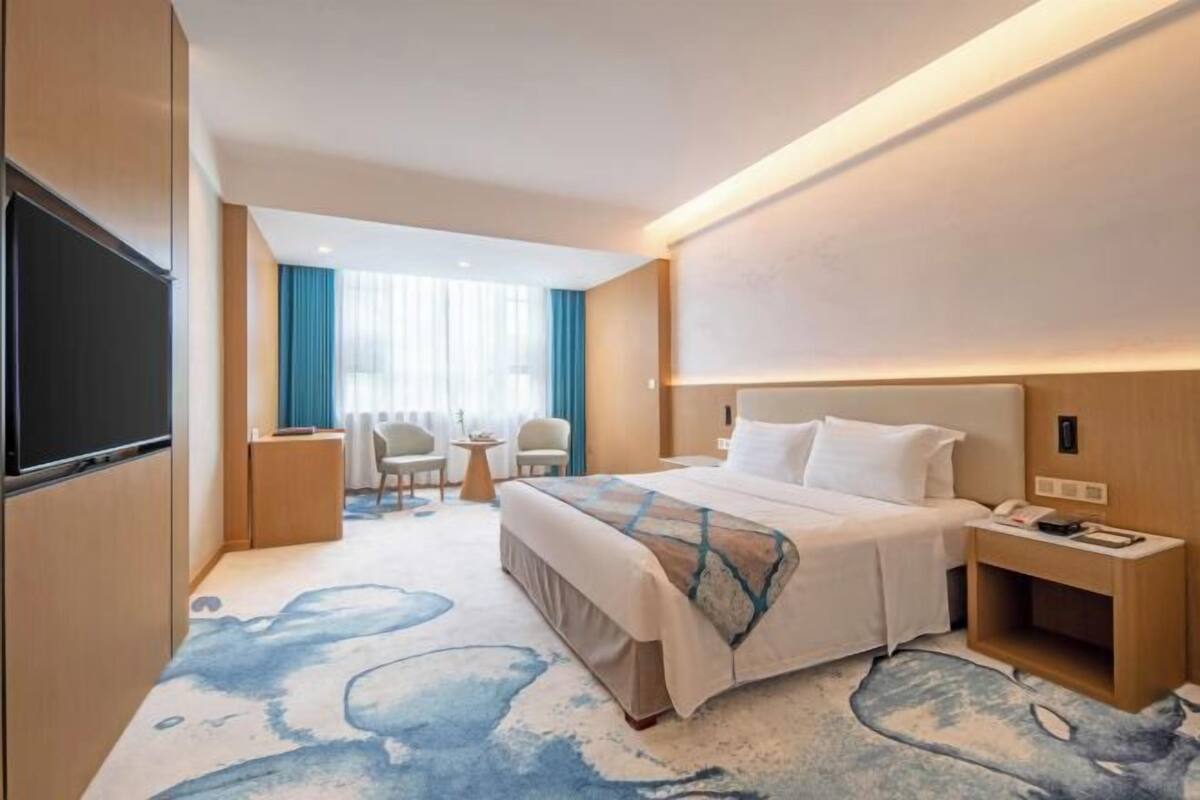 Quarto de hotel com cama, mesa de cabeceira, televisão e janela com cortinas.