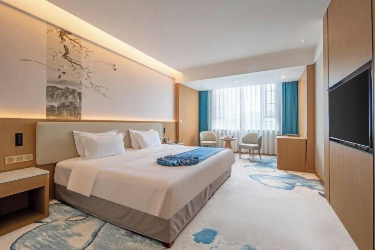 Quarto de hotel com cama, mesa de cabeceira e uma janela grande com cortinas.