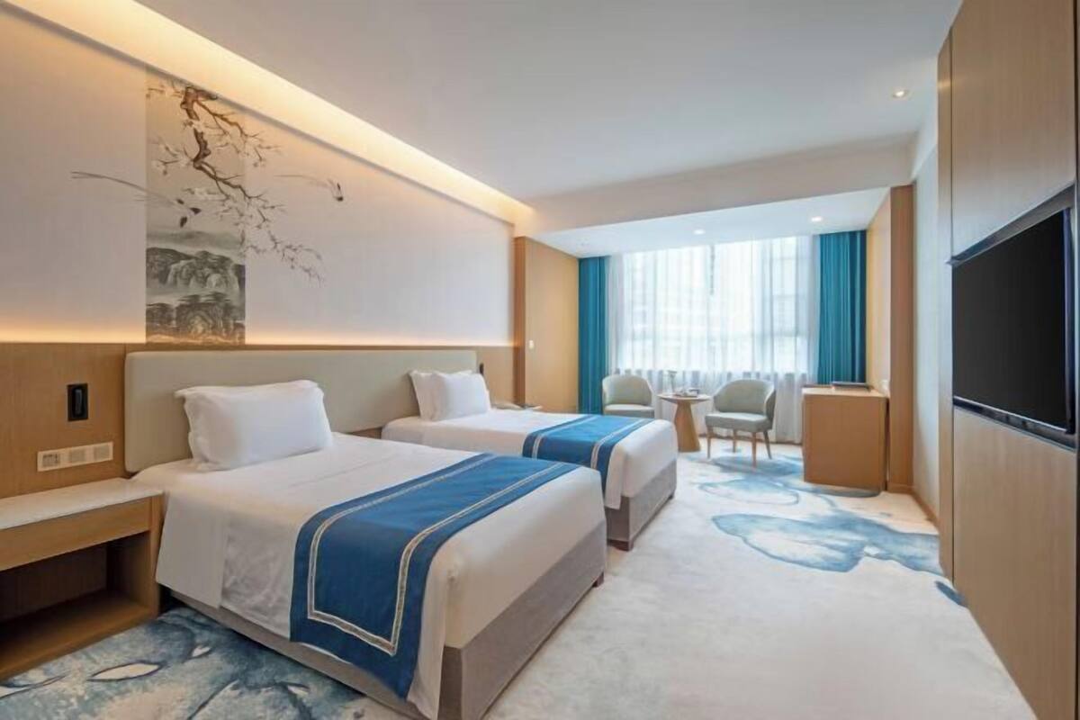 Quarto de hotel com duas camas, uma escrivaninha, uma cadeira, televisão e uma janela grande com cortinas.