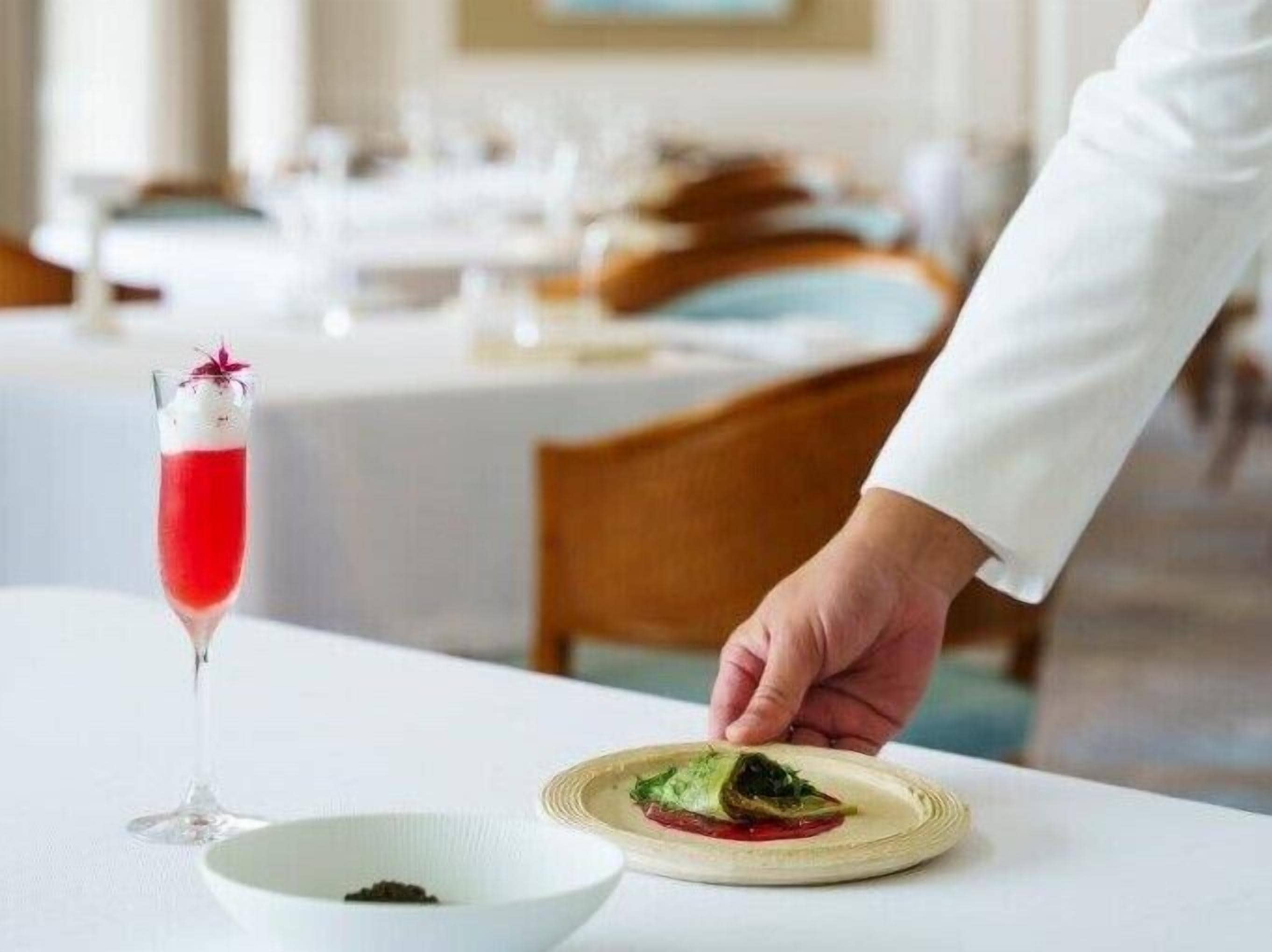 Uma mesa de jantar com um copo de coquetel vermelho, um prato com um acompanhamento de legumes verdes e uma tigela branca com uma substância escura.