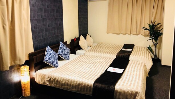 Quarto de hotel com duas camas, cortinas bege, ar condicionado fixado na parede e um vaso de planta.