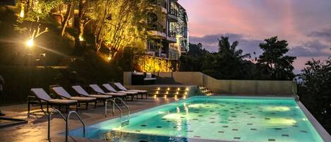 Piscina com espreguiçadeiras e um edifício com janelas iluminadas ao entardecer.